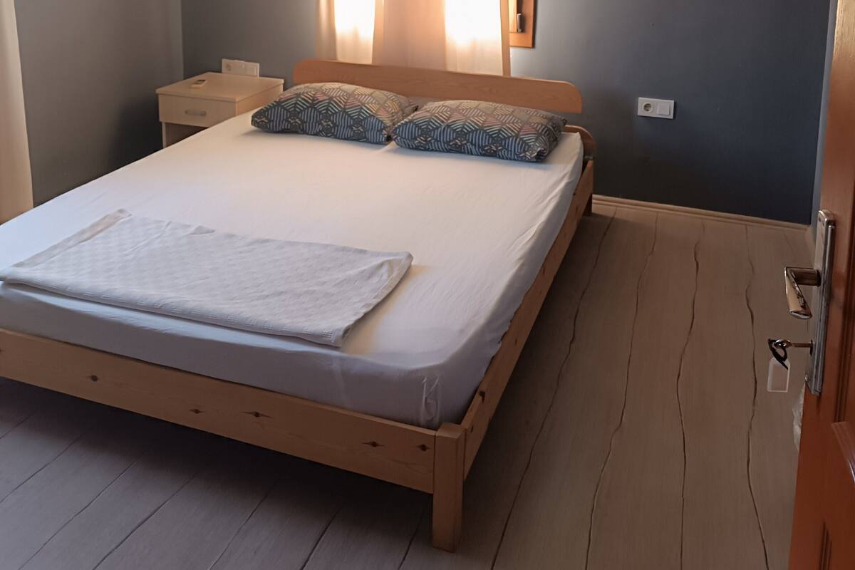 Quarto com cama de madeira, duas mesas de cabeceira, ar condicionado fixado na parede e janela com cortinas leves.
