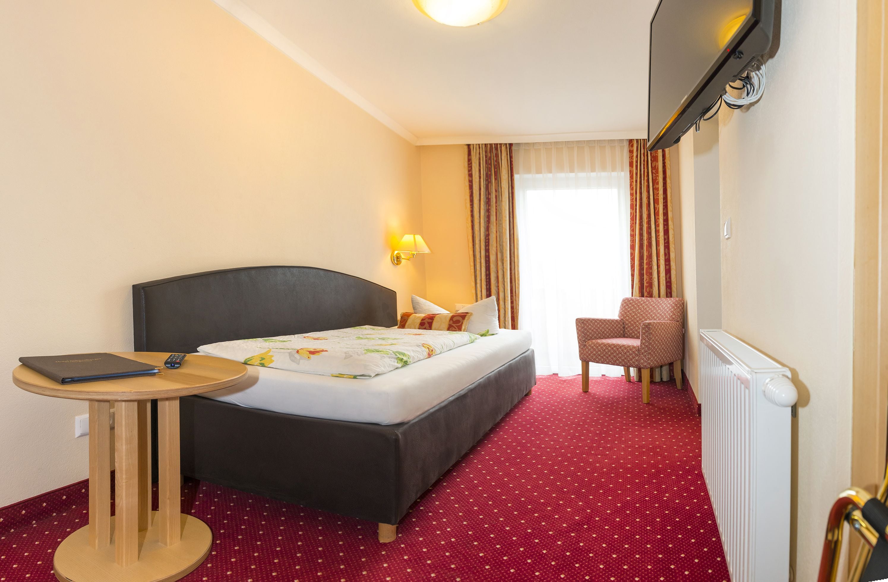 Quarto de hotel com cama, uma mesinha, uma cadeira, televisão fixada na parede e janela com cortinas.