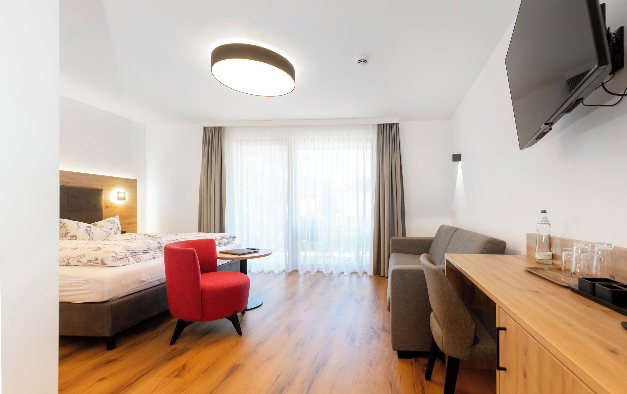 Quarto de hotel com cama, uma cadeira vermelha, um sofá cinza, escrivaninha de madeira e televisão fixada na parede.