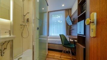 Quarto de hotel com chuveiro em cabine de vidro, cama com vista para o exterior, escrivaninha com cadeira e televisão.