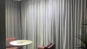 Quarto moderno com uma janela grande coberta por persianas verticais, uma mesa redonda com uma única cadeira e um sofá.