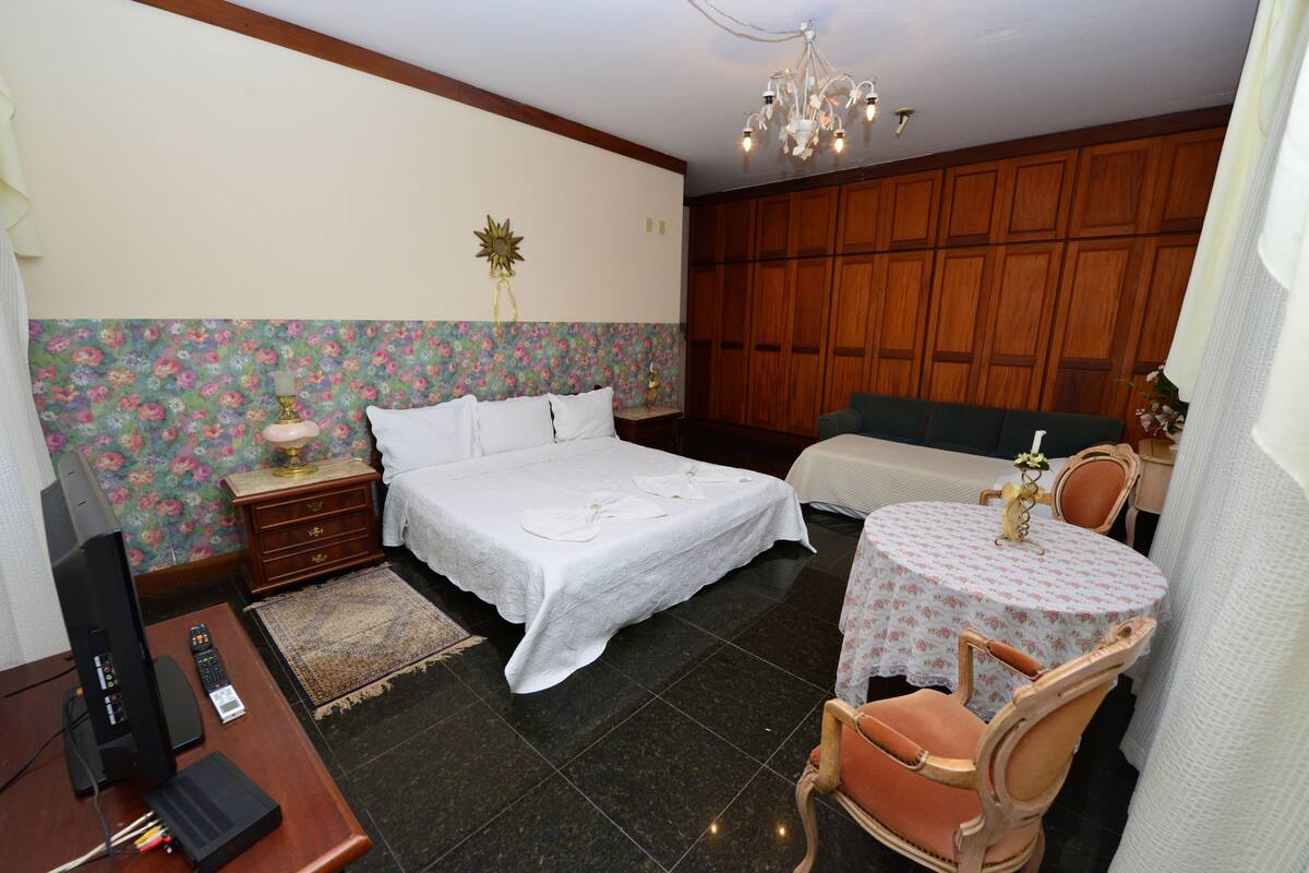 Quarto de hotel com cama, secretária com computador, cadeira, mesa pequena com abajur e um objeto decorativo na parede.