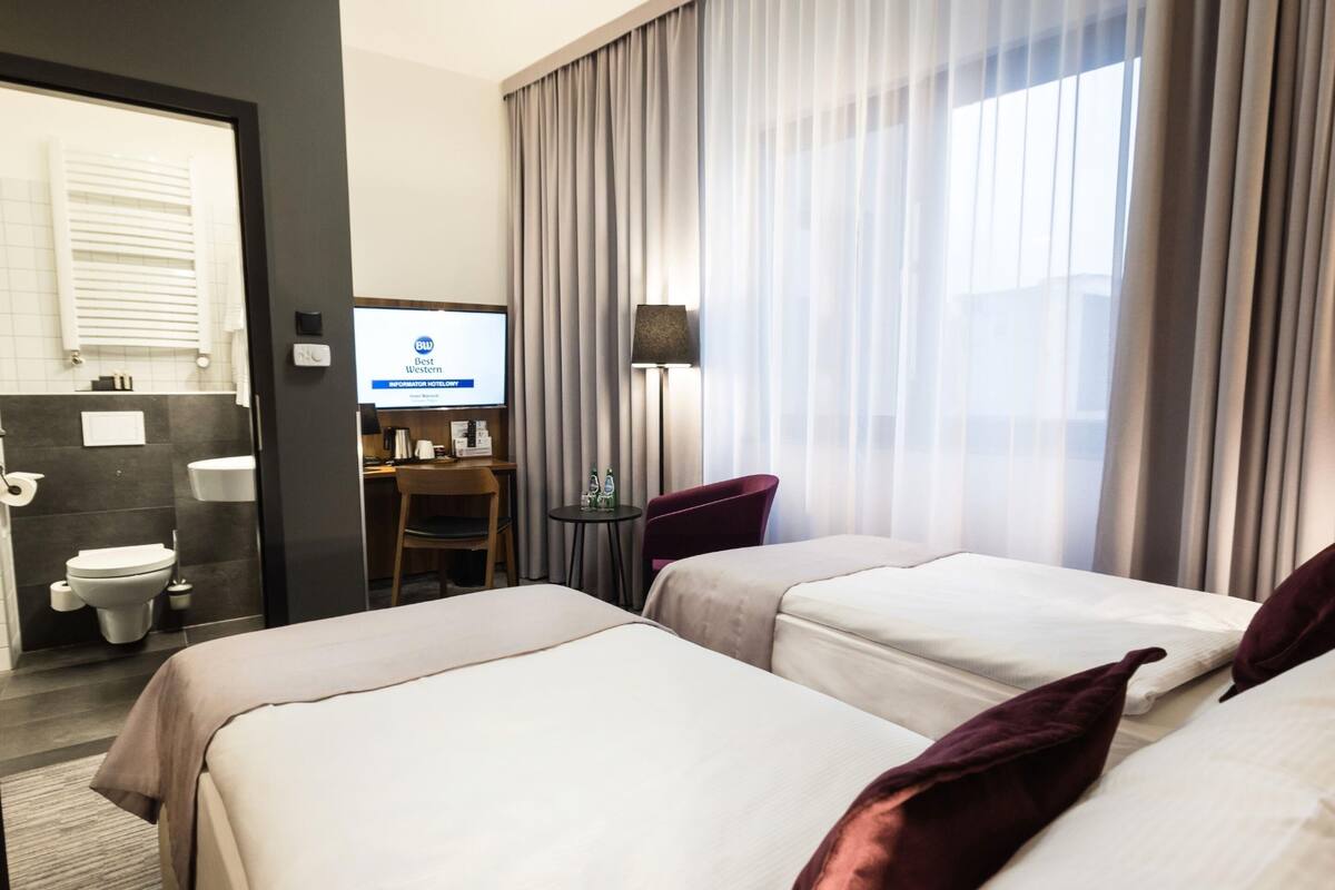 Quarto de hotel com duas camas, uma escrivaninha, uma cadeira, uma televisão e um banheiro.