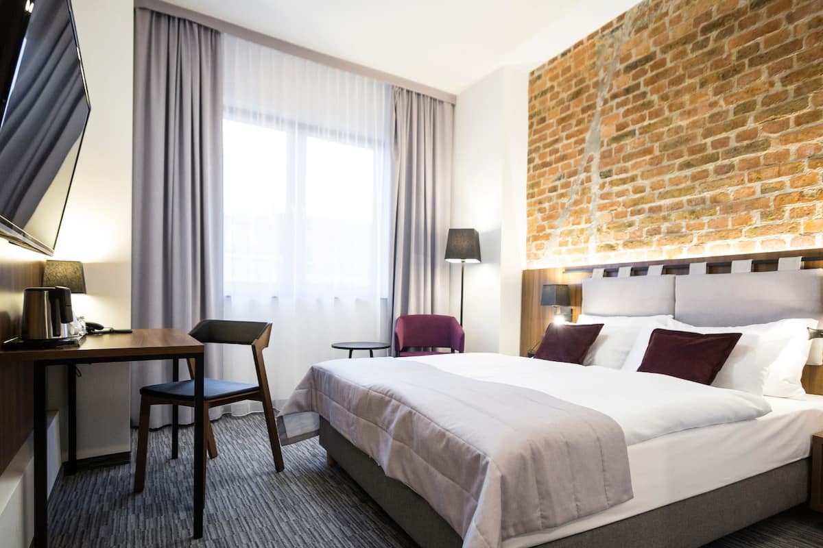 Quarto de hotel com uma cama grande, uma escrivaninha com cadeira, uma mesinha e uma televisão.