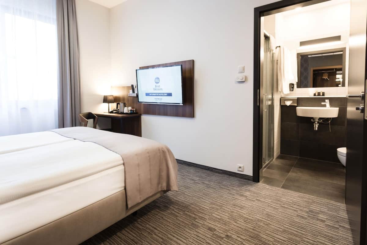 Quarto de hotel com cama, escrivaninha, televisão exibindo 'Best Western' e banheiro com pia e espelho.