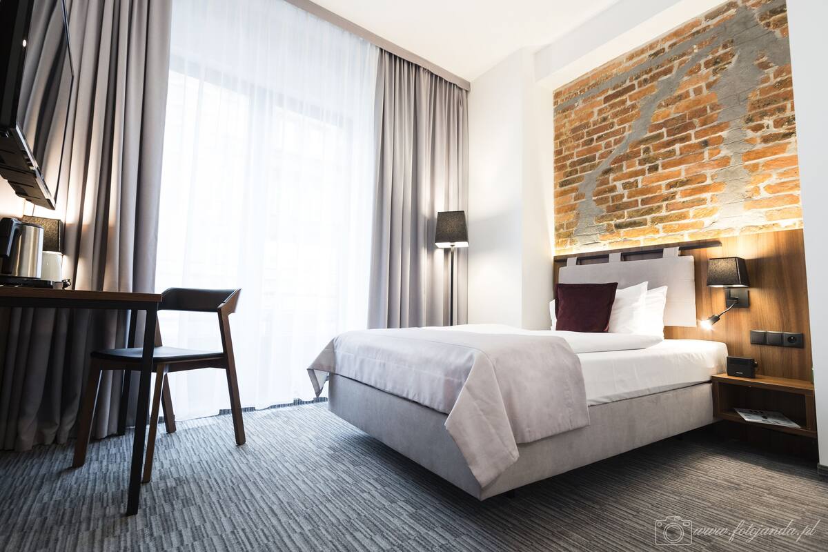 Quarto de hotel com cama, escrivaninha, cadeira, luminária e parede com padrão de tijolos.