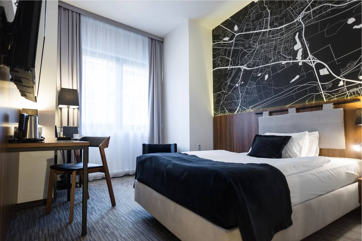 Quarto de hotel com cama, escrivaninha e cadeira, luminária, cafeteira e um mapa de parede grande.