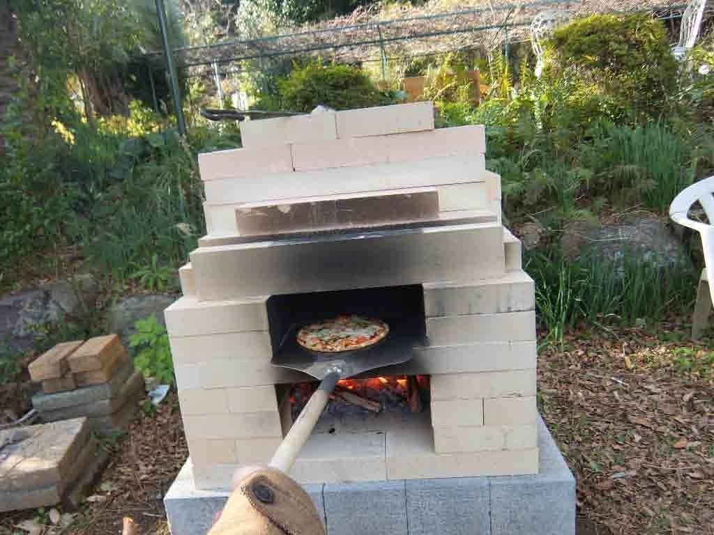 Forno a lenha de tijolos ao ar livre, com uma pizza dentro, rodeado por vegetação.