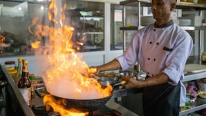 Um cozinheiro preparando comida em uma wok grande sobre um fogão, com chamas visíveis, em uma cozinha.