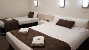 Duas camas de solteiro com cabeceiras marrons e roupa de cama branca, uma mesinha de cabeceira com telefone e despertador, e um candeeiro fixo na parede.
