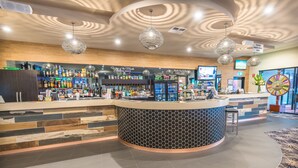 Um saguão de hotel com uma recepção central, diversas garrafas de bebidas alcoólicas e uma grande roda decorativa.