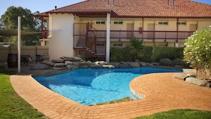Uma piscina com deck de tijolos, um alpendre coberto e um edifício com telhado de telhas vermelhas ao fundo.
