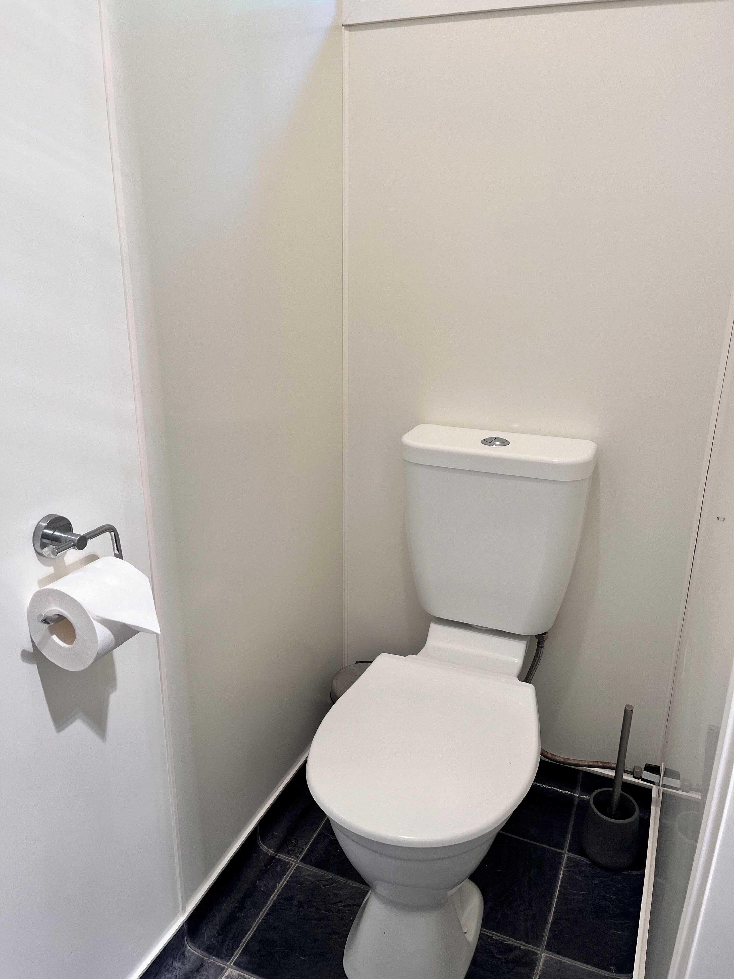 Uma sanita pequena e branca, com botão de descarga e porta-papel higiénico na parede.