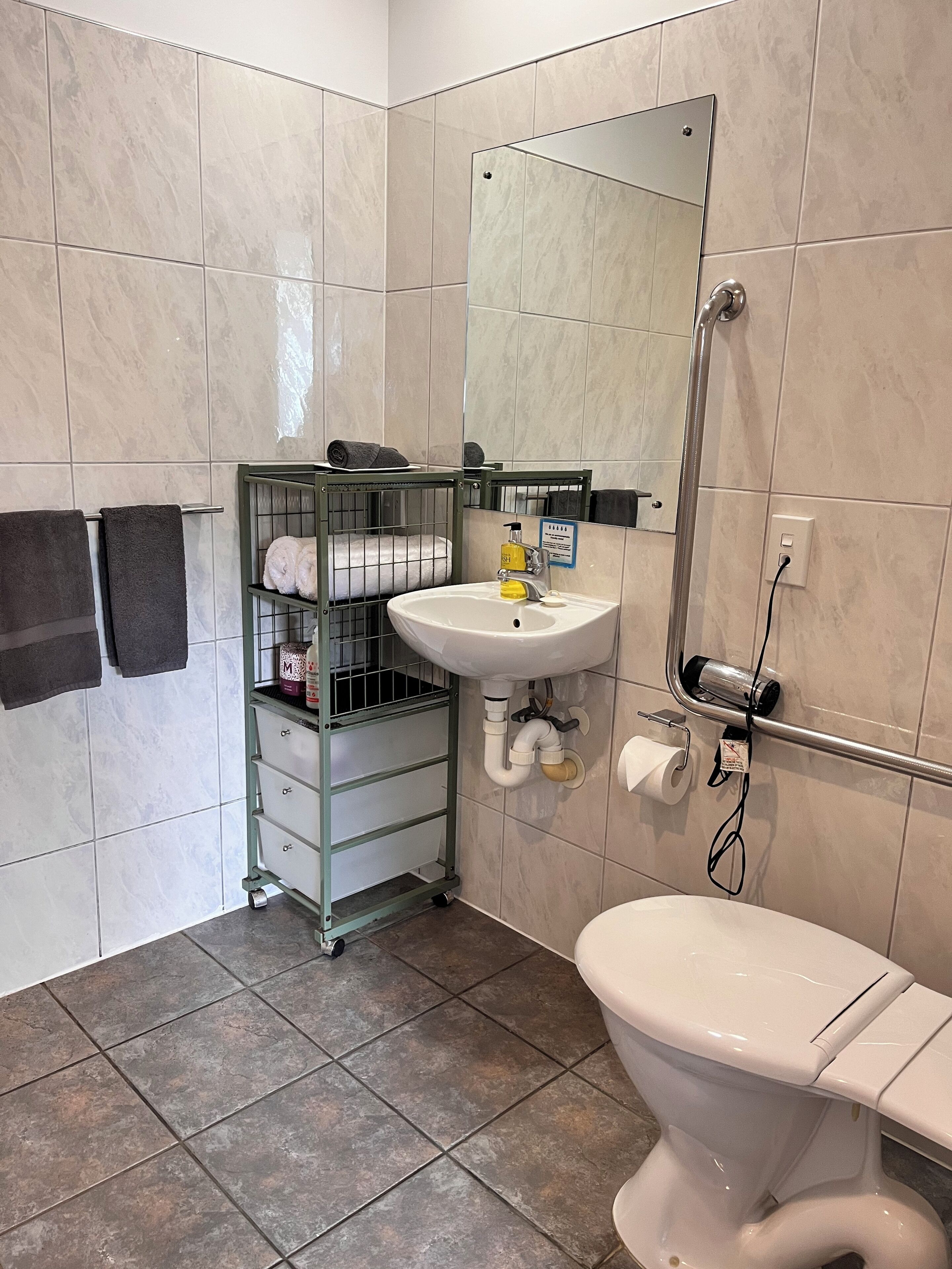 Casa de banho com lavatório branco, sanita e área de chuveiro. Tem um porta-toalhas com toalhas e uma estante metálica com artigos de higiene pessoal.