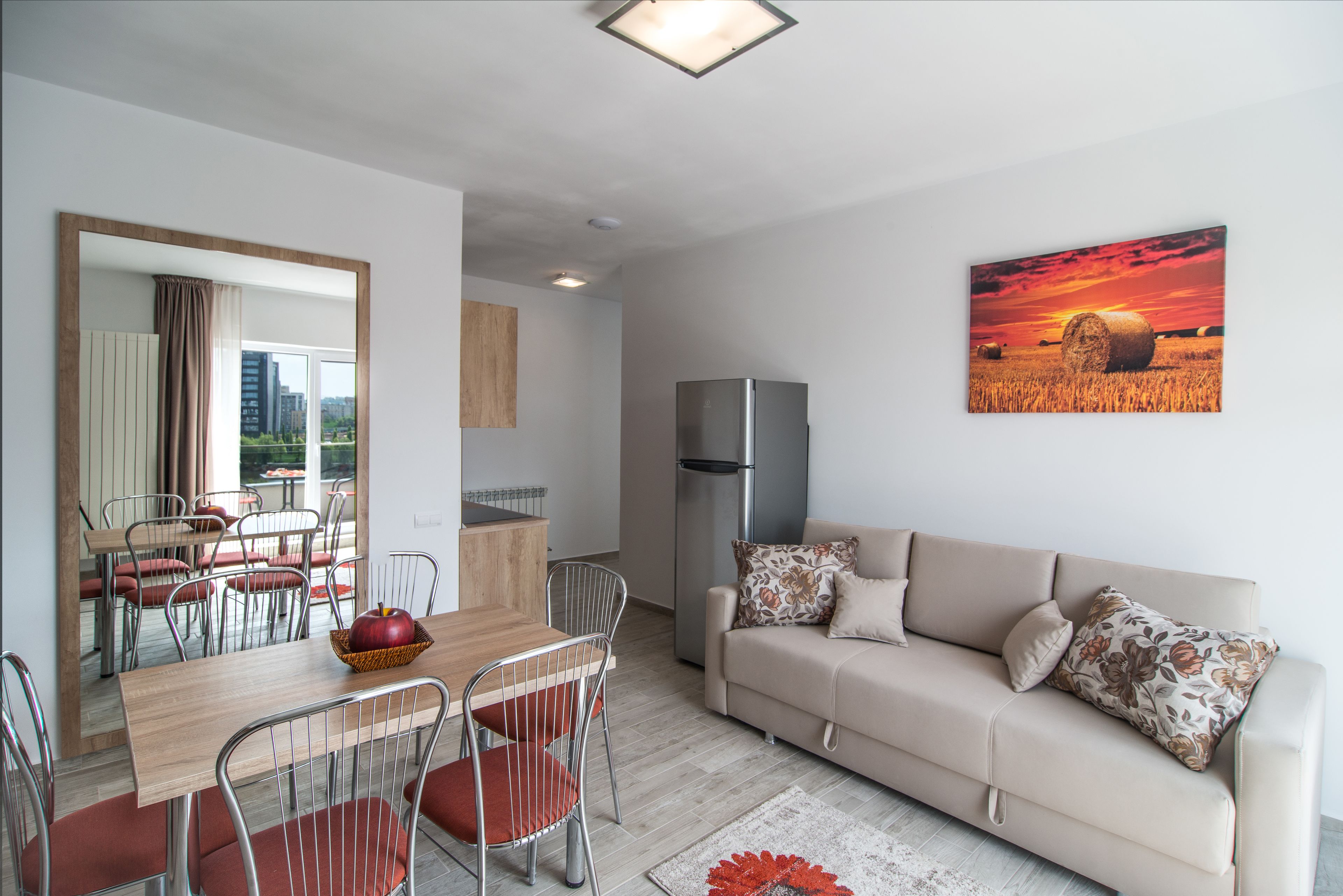 Uma sala de estar moderna com um sofá, mesa de jantar, cadeiras e uma grande pintura de um pôr do sol sobre um campo.