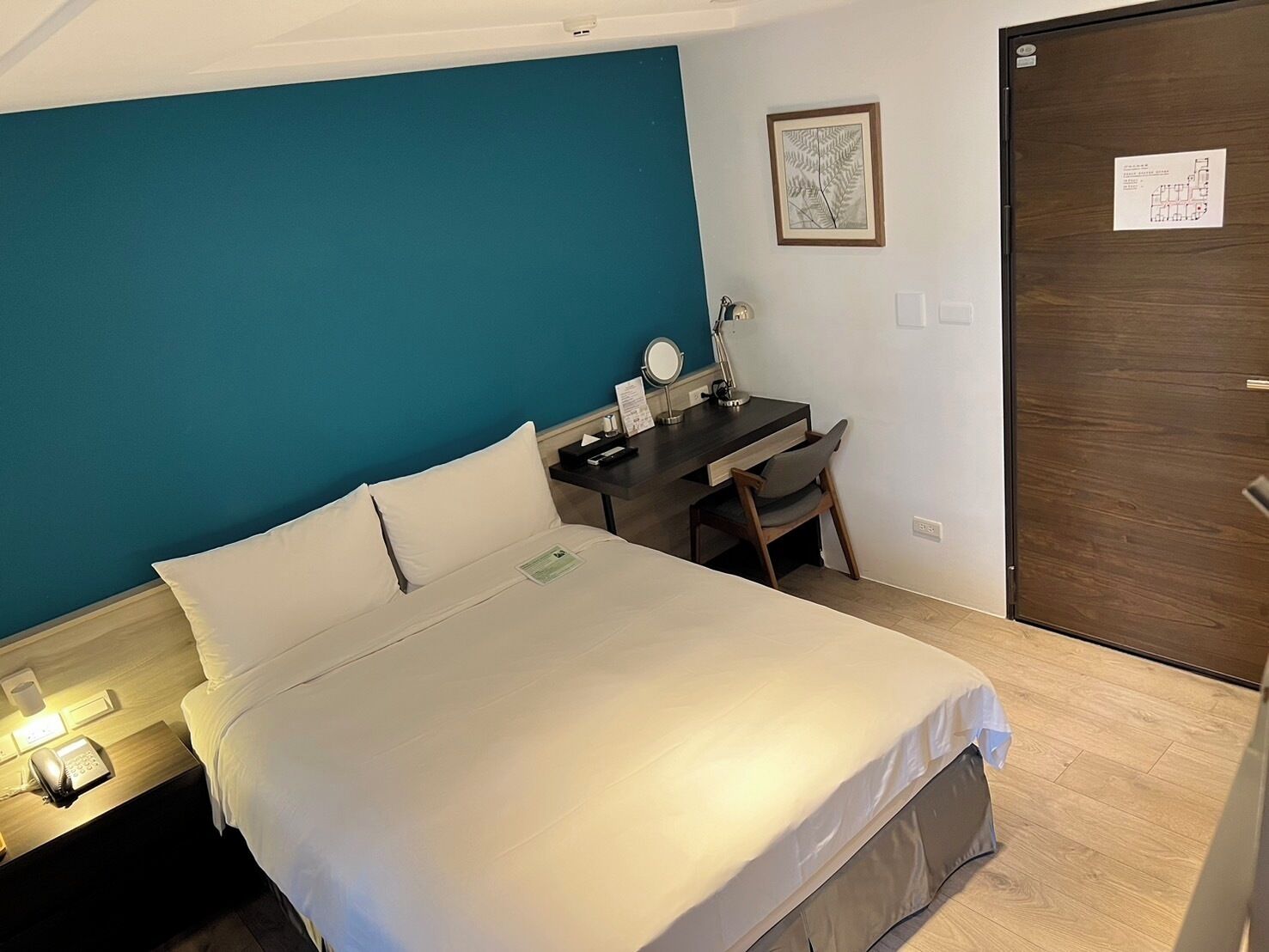 Quarto de hotel com cama, escrivaninha, cadeira e porta.