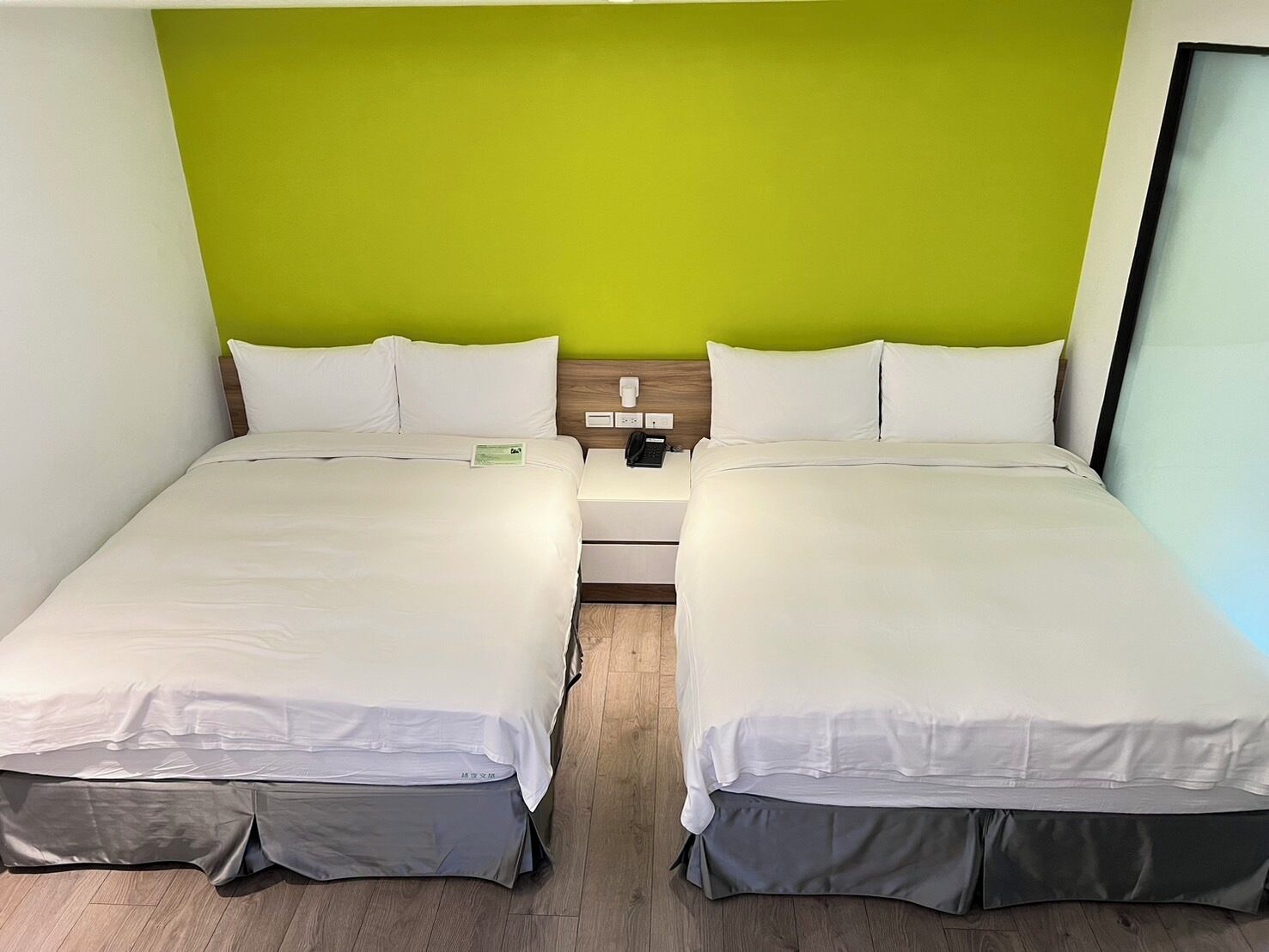 Quarto duplo com duas camas, mesa de cabeceira com telefone e parede verde.