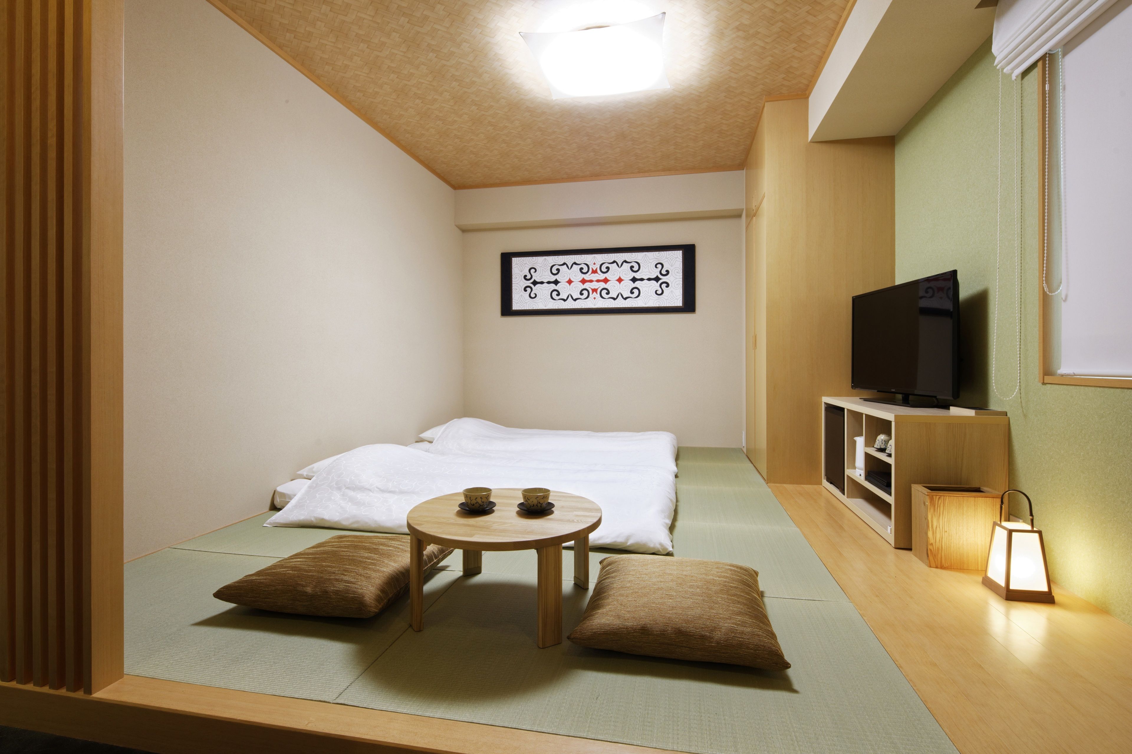 Um quarto tradicional japonês com tatames, uma mesa de madeira baixa com duas xícaras, uma televisão sobre um suporte de madeira e um quadro pendurado na parede.