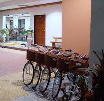 Uma fileira de bicicletas com cestas, estacionadas em piso de azulejos em área pública, com uma porta de madeira e uma divisória de vidro ao fundo.
