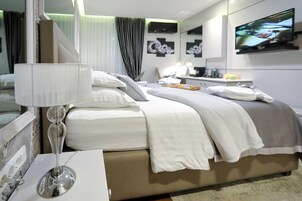 Quarto de hotel moderno com uma cama grande, uma mesa de cabeceira com abajur, uma TV na parede e uma pequena escrivaninha com cadeira.