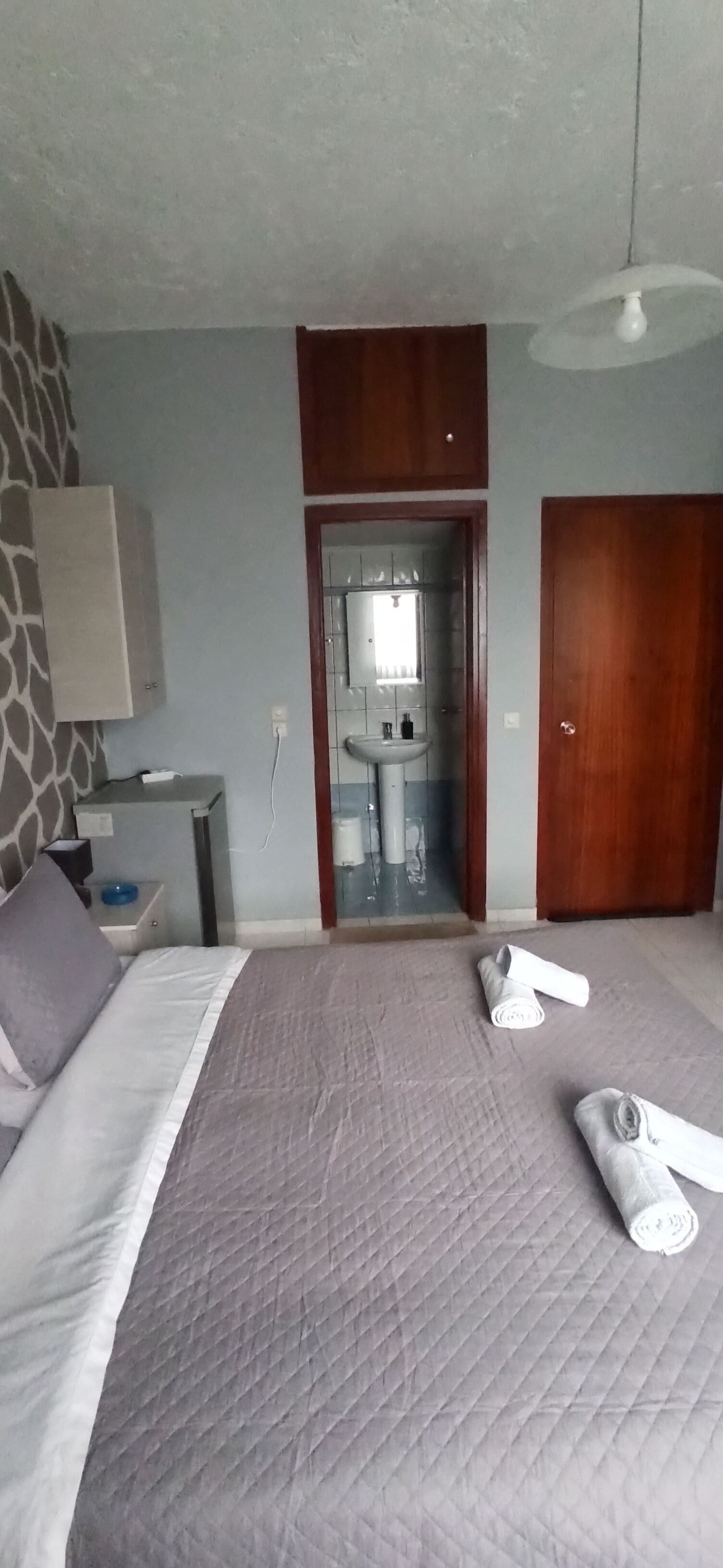 Quarto de hotel com cama, mesa de cabeceira, espelho, guarda-roupa e banheiro visível através de uma porta aberta.