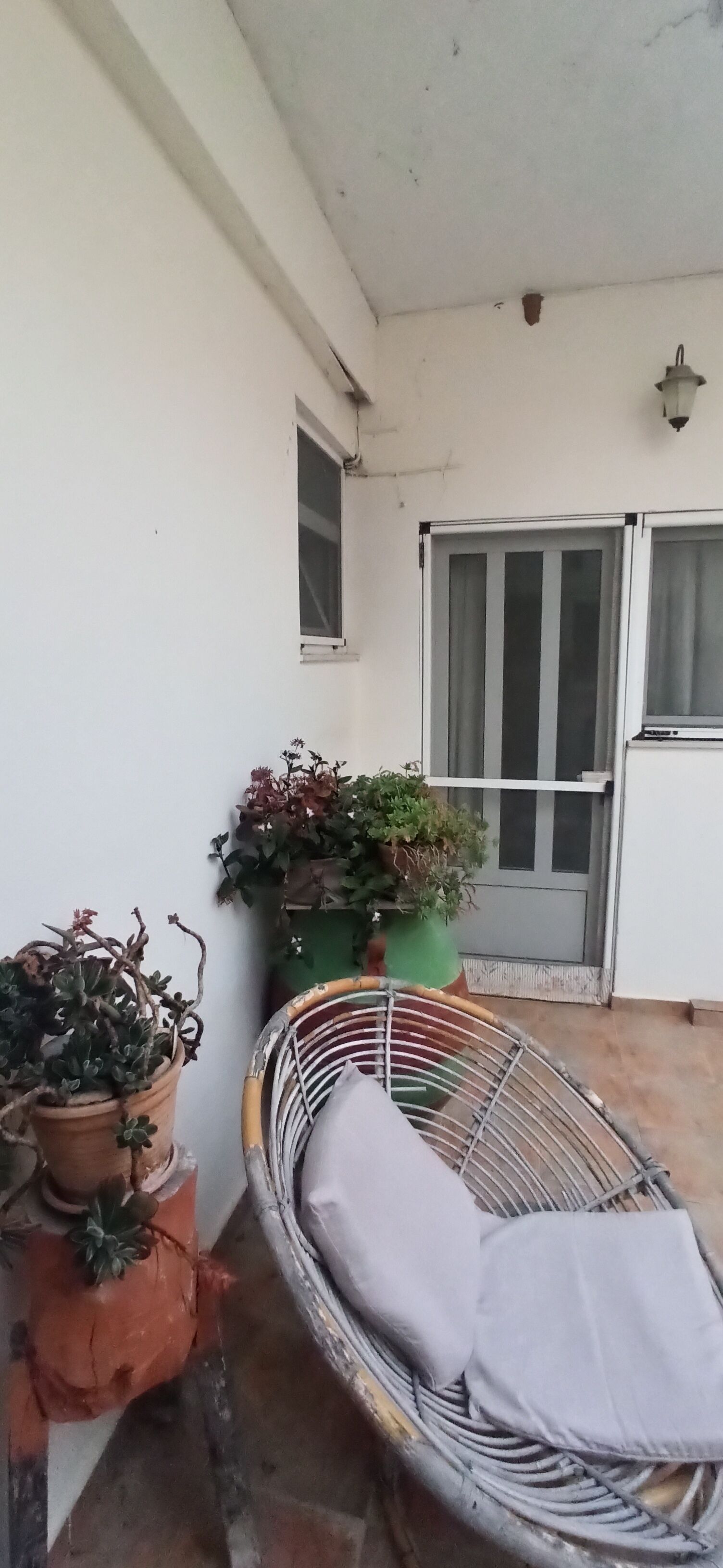 Uma varanda com cadeira de vime, plantas em vasos e porta de correr de vidro.