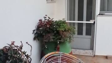 Uma varanda com cadeira de vime, plantas em vasos e porta de correr de vidro.