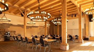 Uma sala de conferências ampla, com vigas de madeira, várias luminárias pendentes e parede de tijolos. Mesas e cadeiras dispostas para reuniões.