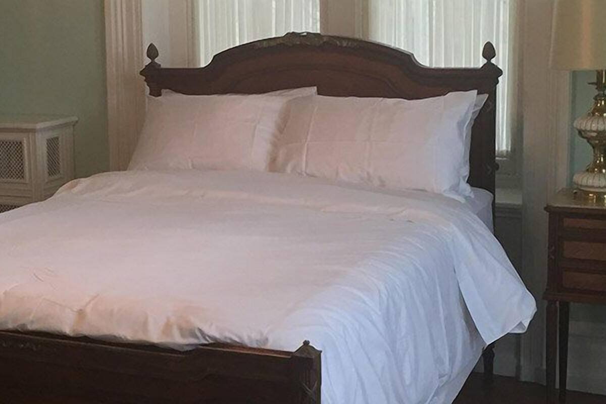Uma cama bem arrumada com cabeceira de madeira, mesa de cabeceira com abajur e um pequeno criado-mudo branco com caixa de lenços.
