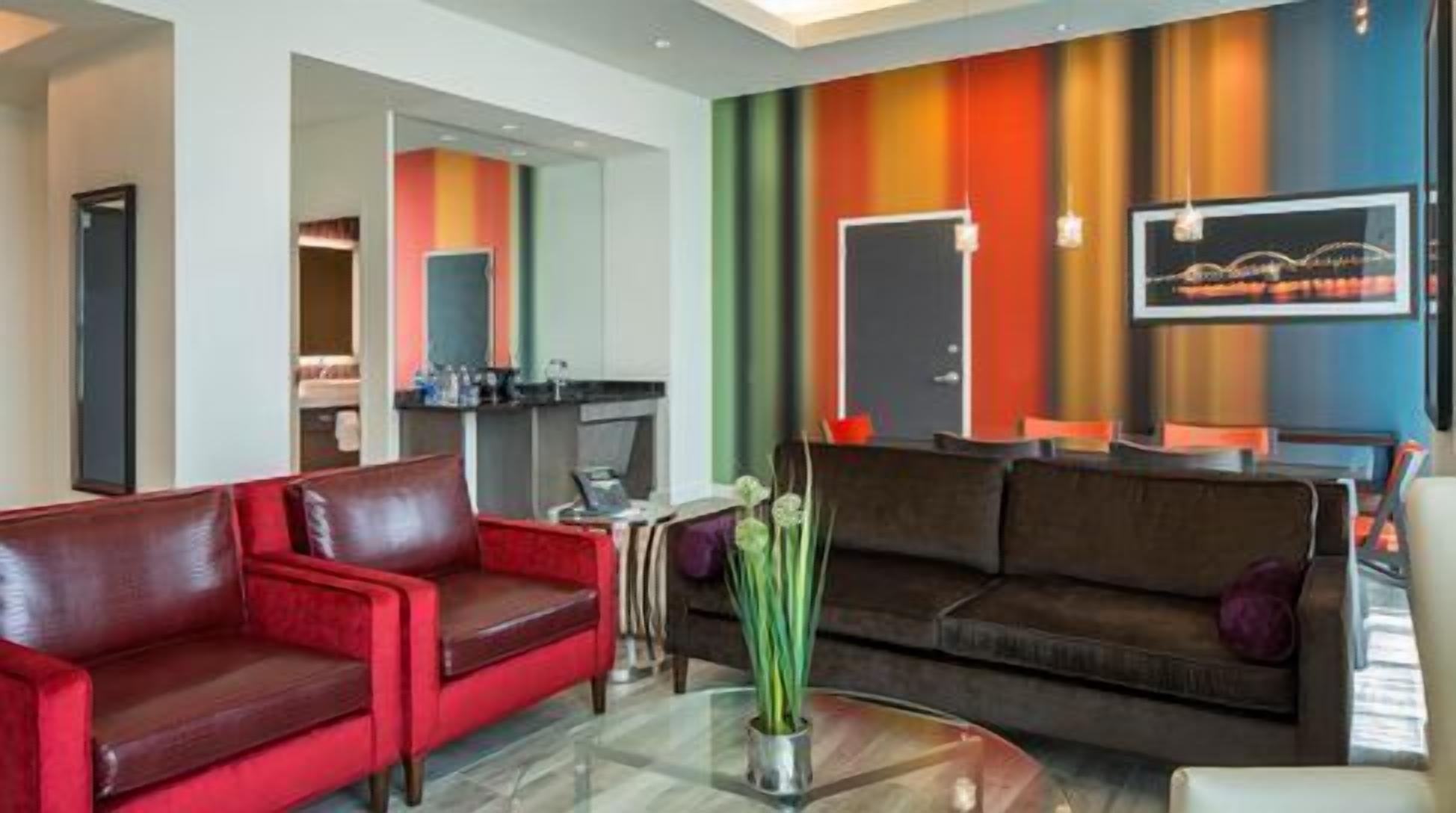 Um hall de hotel com um sofá vermelho, um sofá marrom, uma mesa de centro de vidro e uma parede colorida.