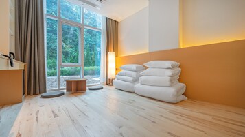 Quarto com uma janela ampla, cama com travesseiros brancos, luminária de pé e uma pequena mesa de madeira.