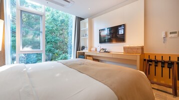 Quarto de hotel com uma cama grande, uma escrivaninha com computador, televisão e vista para o verde lá fora.