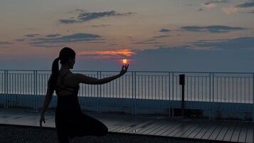 Uma pessoa em um deck de madeira com guarda-corpo de metal, de frente para o pôr do sol.