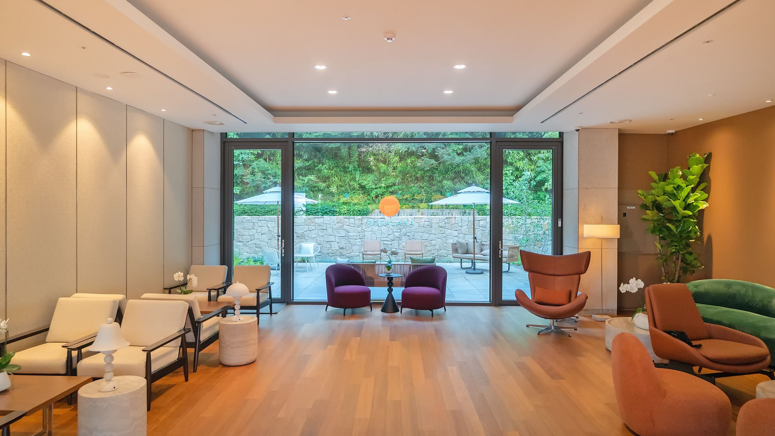 Um interior moderno com piso de madeira, área de estar com cadeiras e mesas, e vista para um jardim exterior através de grandes portas de vidro.