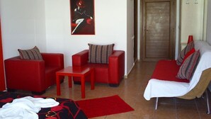 Uma sala de estar moderna com sofás vermelhos, uma mesa pequena e uma cadeira branca. Há uma porta ao fundo e um tapete vermelho no chão.