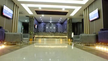 Um lobby moderno com balcão de recepção em mármore, área de estar e duas televisões de tela plana fixadas nas paredes.