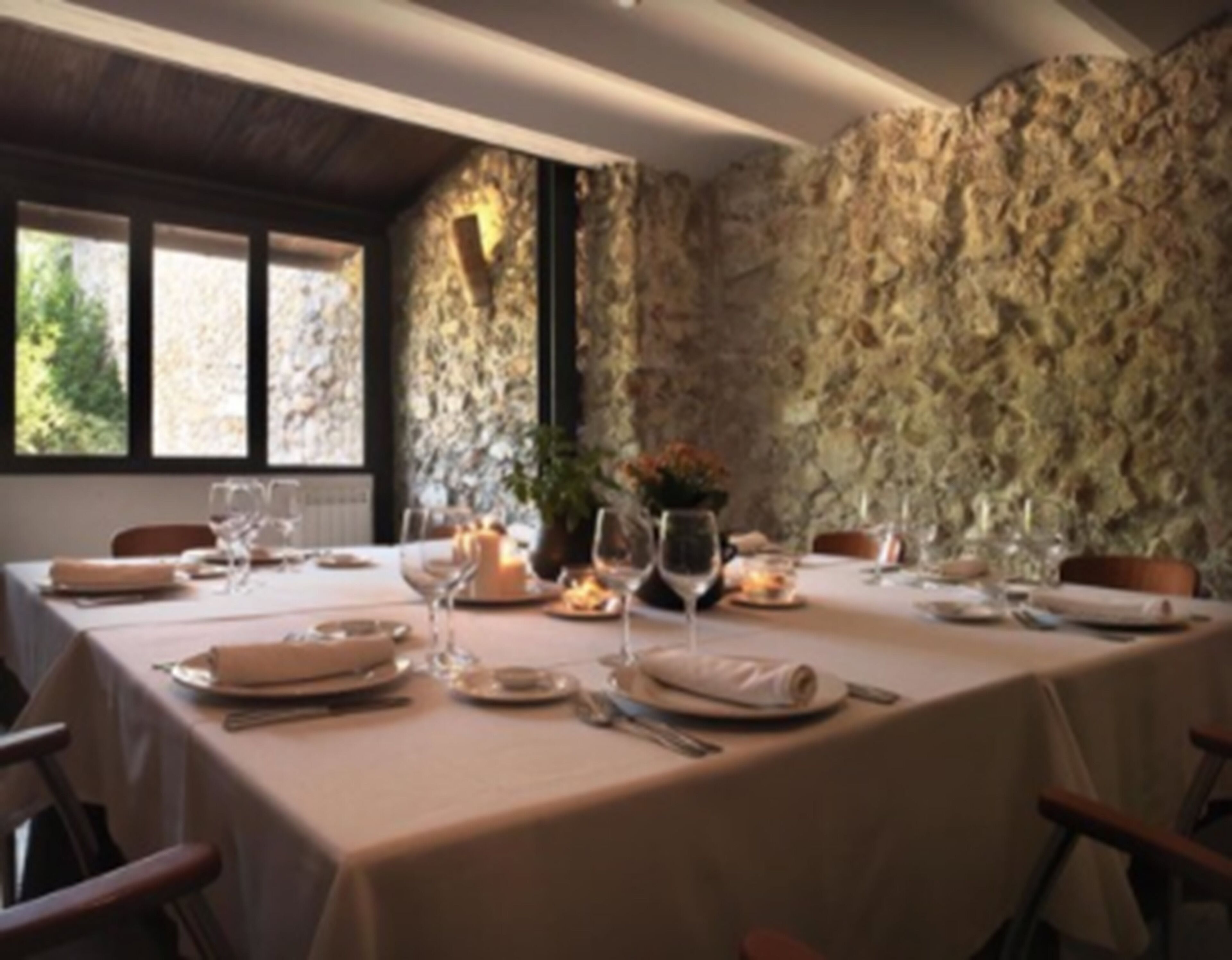 Mesa de jantar posta com pratos, copos e velas, com parede de pedra e janelas ao fundo.