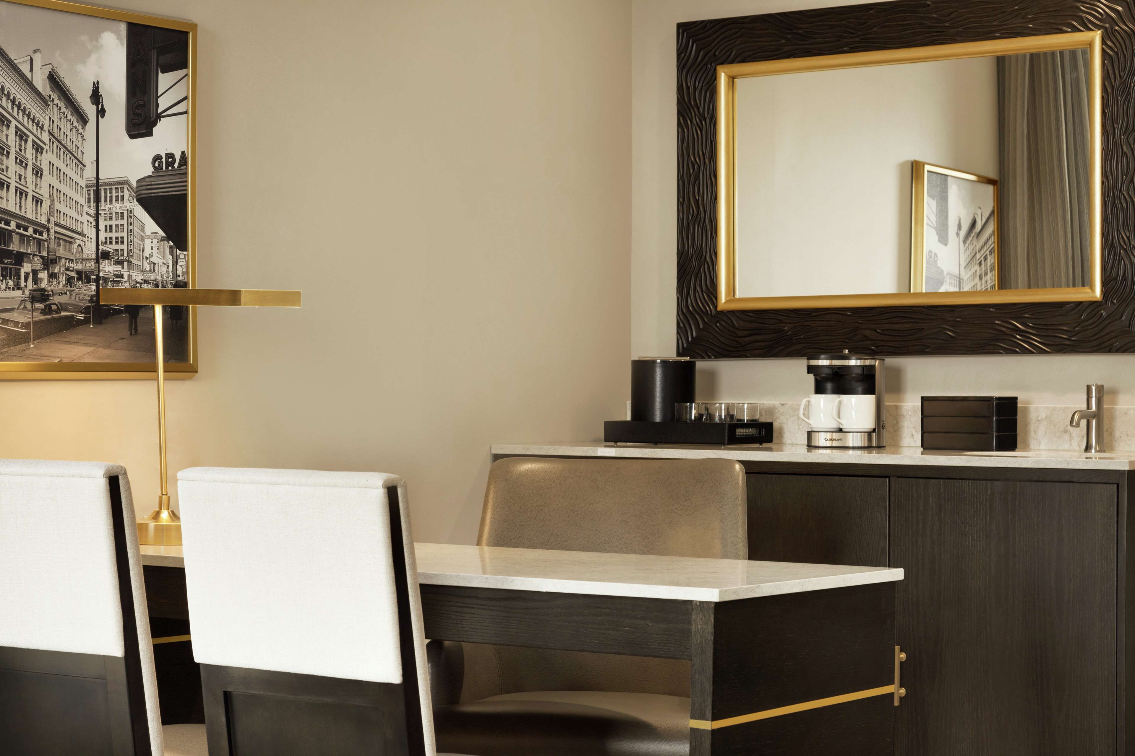 Quarto de hotel moderno com área de jantar, incluindo uma mesa, cadeiras, máquina de café e uma imagem emoldurada da paisagem urbana na parede.