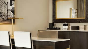 Quarto de hotel moderno com área de jantar, incluindo uma mesa, cadeiras, máquina de café e uma imagem emoldurada da paisagem urbana na parede.