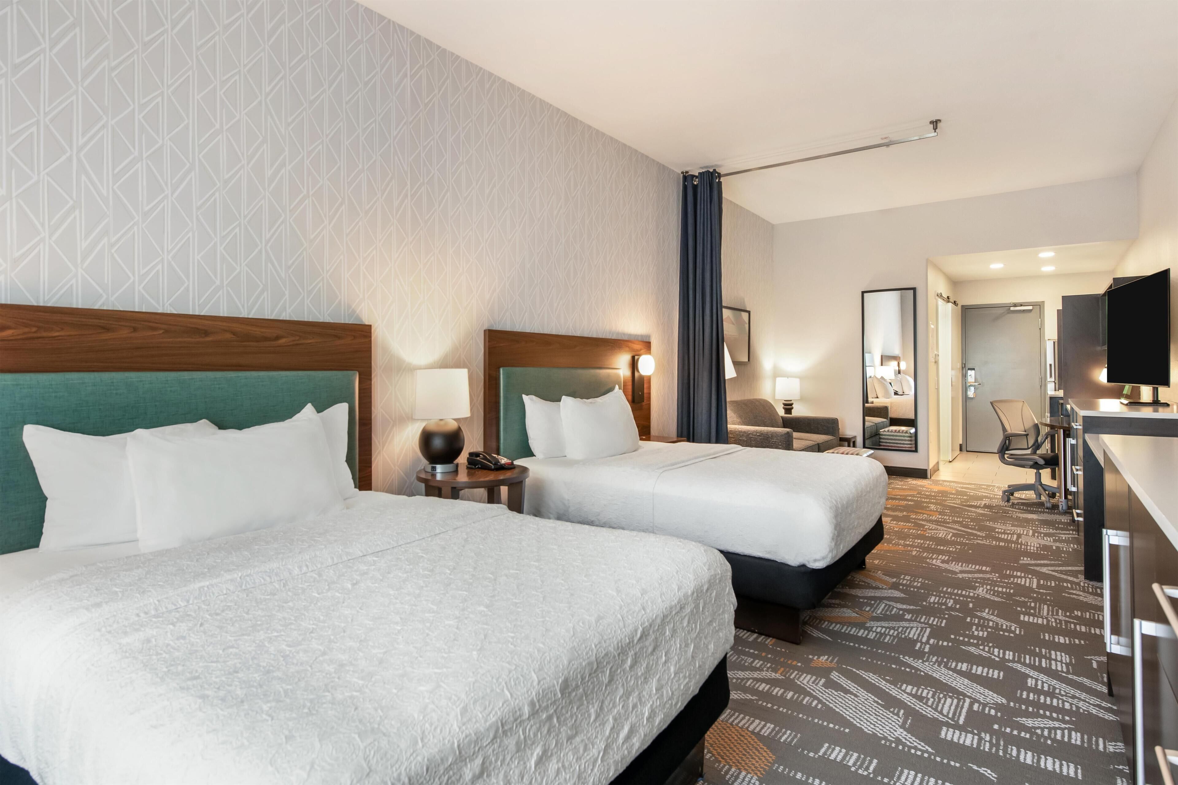 Quarto de hotel com duas camas, uma escrivaninha, uma cadeira, televisão e banheiro visível por uma porta aberta.