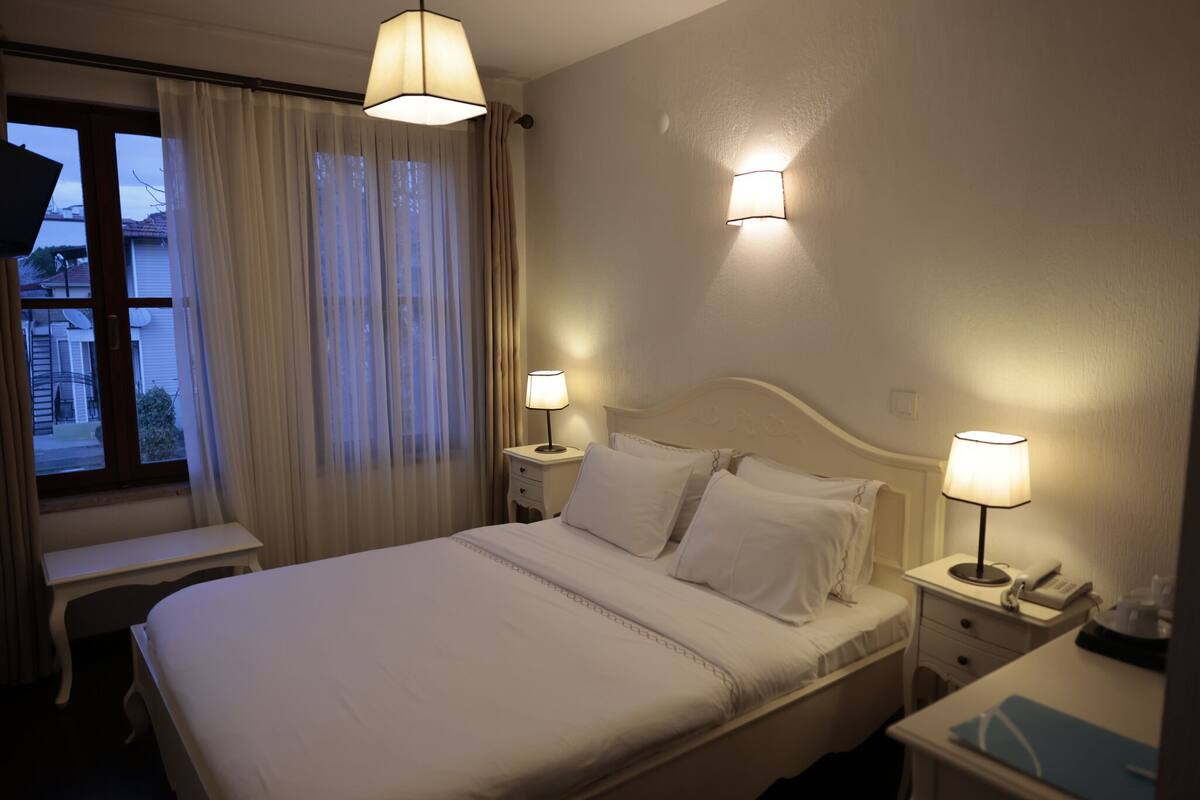 Quarto de hotel com cama, mesas de cabeceira, luminárias, janela com cortinas e vista para o exterior.