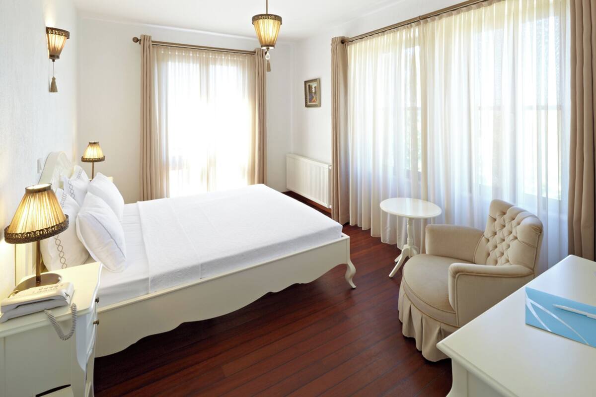 Quarto de hotel com uma cama grande, luminárias de cabeceira, uma cadeira, uma mesinha e uma janela com cortinas.