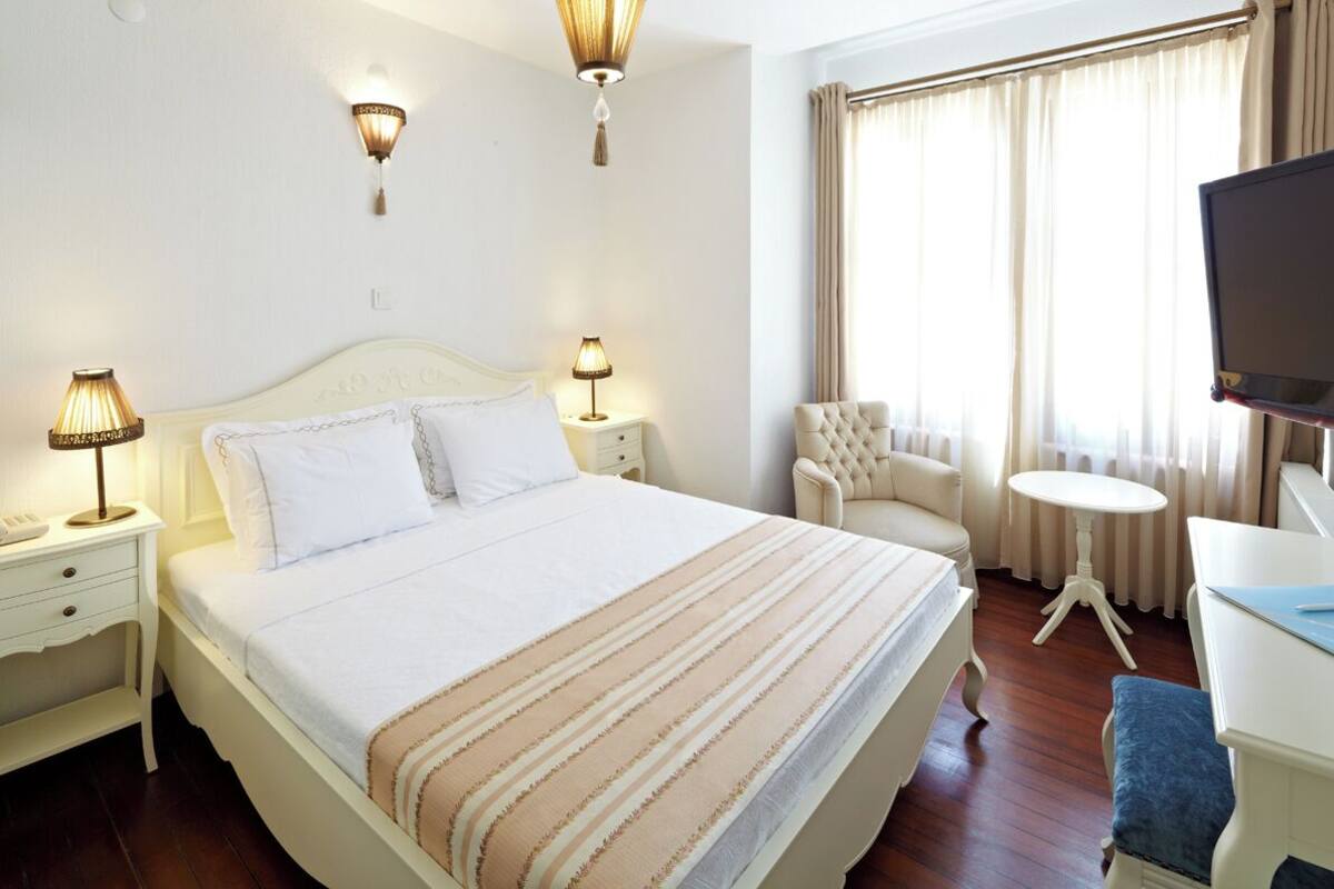 Quarto de hotel com uma cama grande, luminárias de cabeceira, uma cadeira, uma mesa redonda pequena e uma televisão.