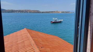 Vista da janela com mar calmo, um barco ao longe e um telhado de azulejos em primeiro plano.