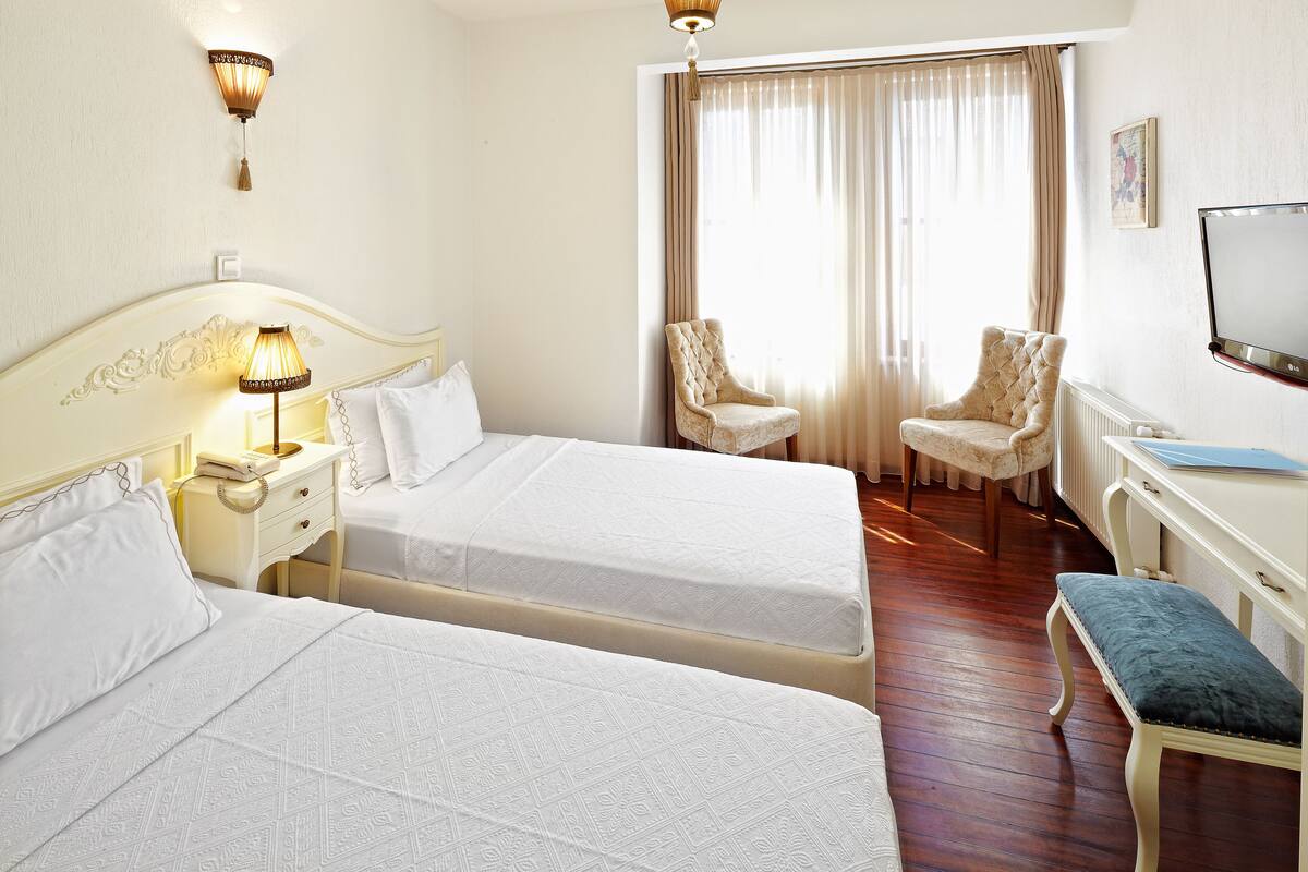 Quarto de hotel com duas camas, área de estar com uma poltrona, escrivaninha e televisão.