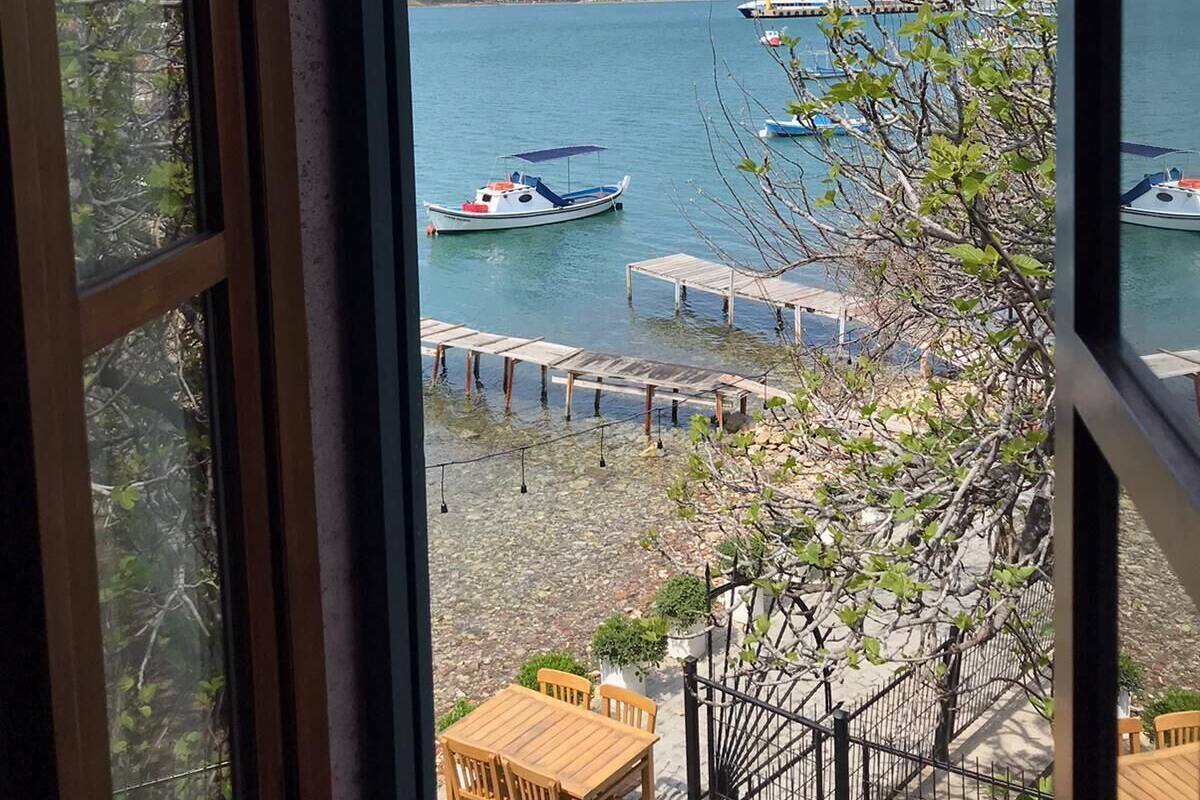 Vista da janela para um corpo d'água calmo, com barcos, um cais e uma pequena mesa e cadeiras de madeira.