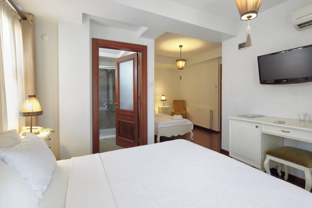 Quarto de hotel com cama, escrivaninha, cadeira, televisão e banheiro visível através de uma porta aberta.