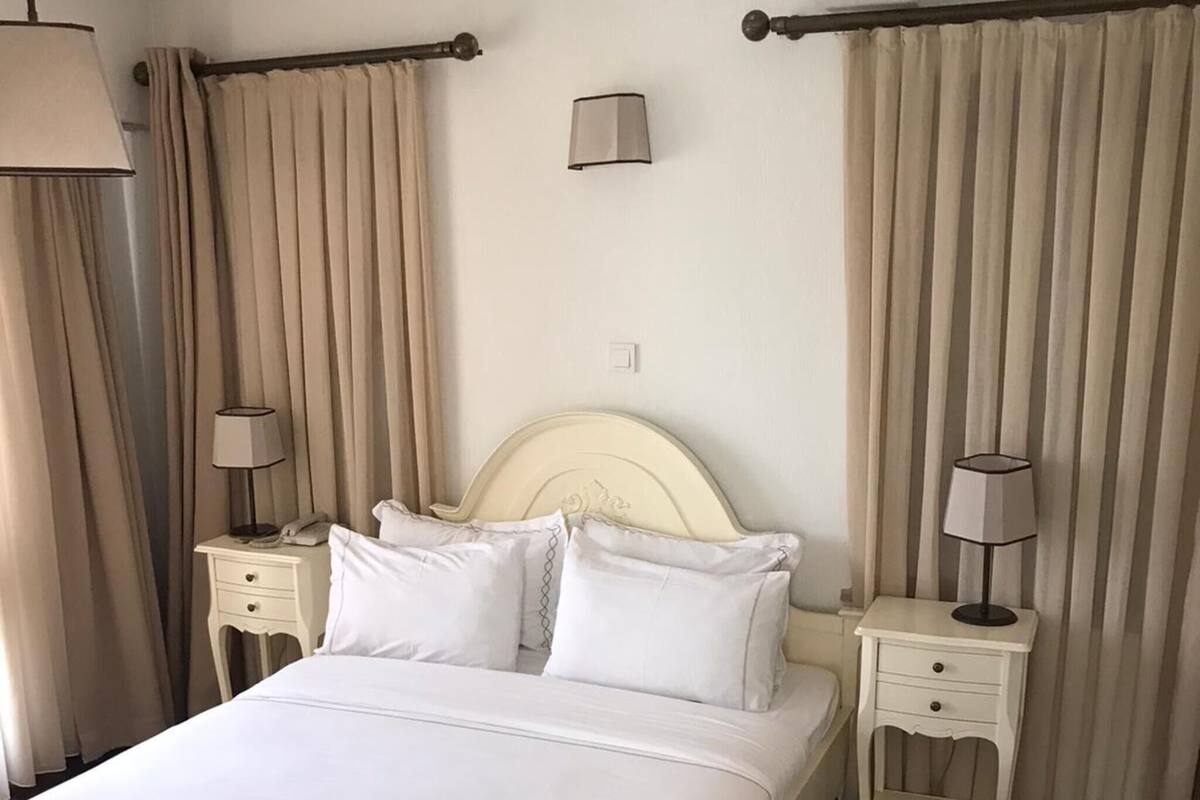 Uma cama bem arrumada com lençóis brancos, duas mesas de cabeceira com abajures e cortinas bege.