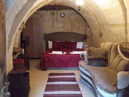 Um quarto aconchegante com um arco de pedra, uma cama com colcha vermelha, uma mesinha de cabeceira de madeira, um sofá de couro e um tapete listrado.