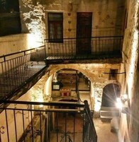 Uma passagem estreita e pouco iluminada, com parede de pedra e grades de metal, que leva a um pequeno pátio com um banco e uma porta.